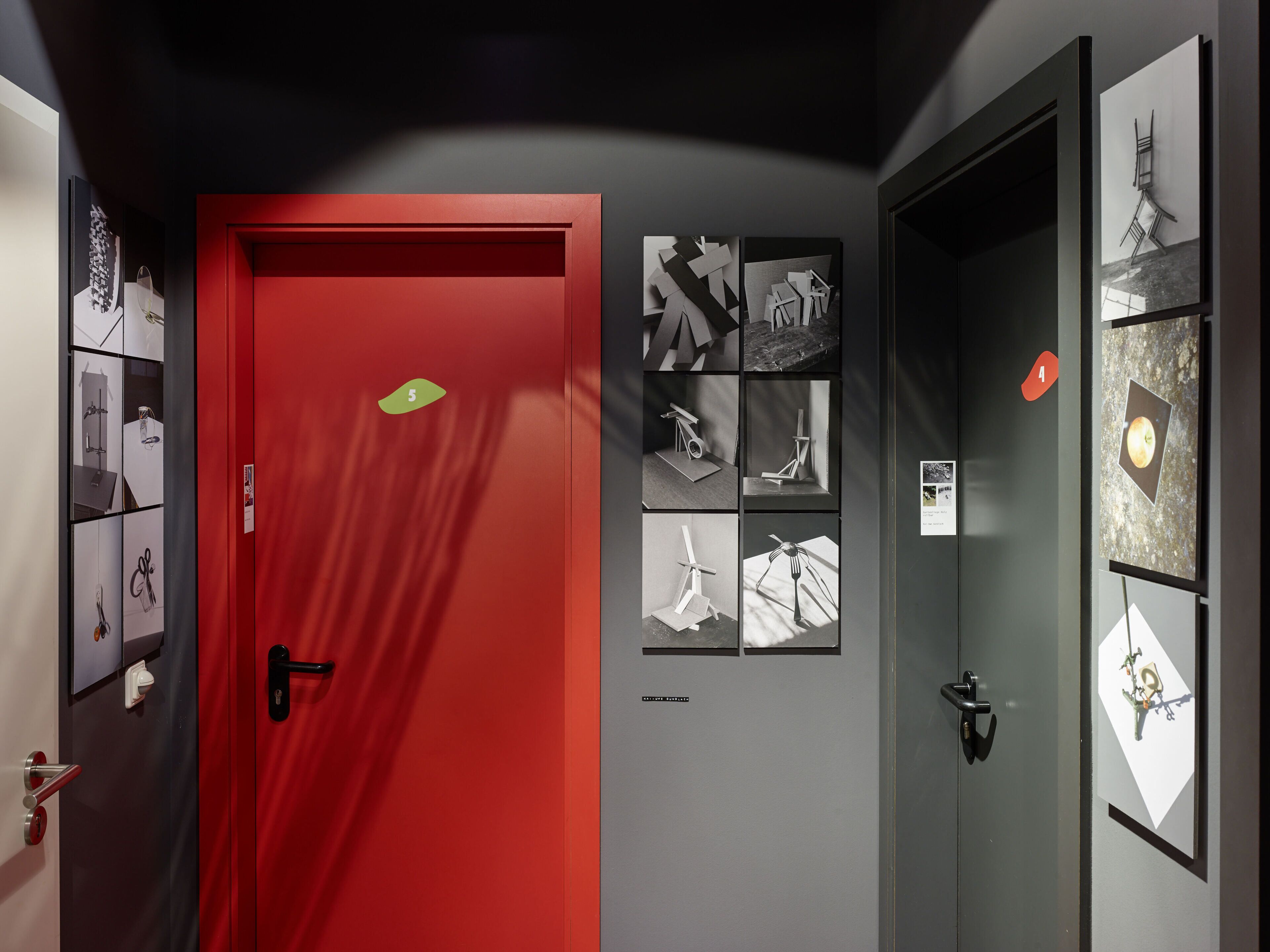 Um corredor com uma porta vermelha, uma série de fotografias preto e branco emolduradas nas paredes e uma placa com o número da porta.