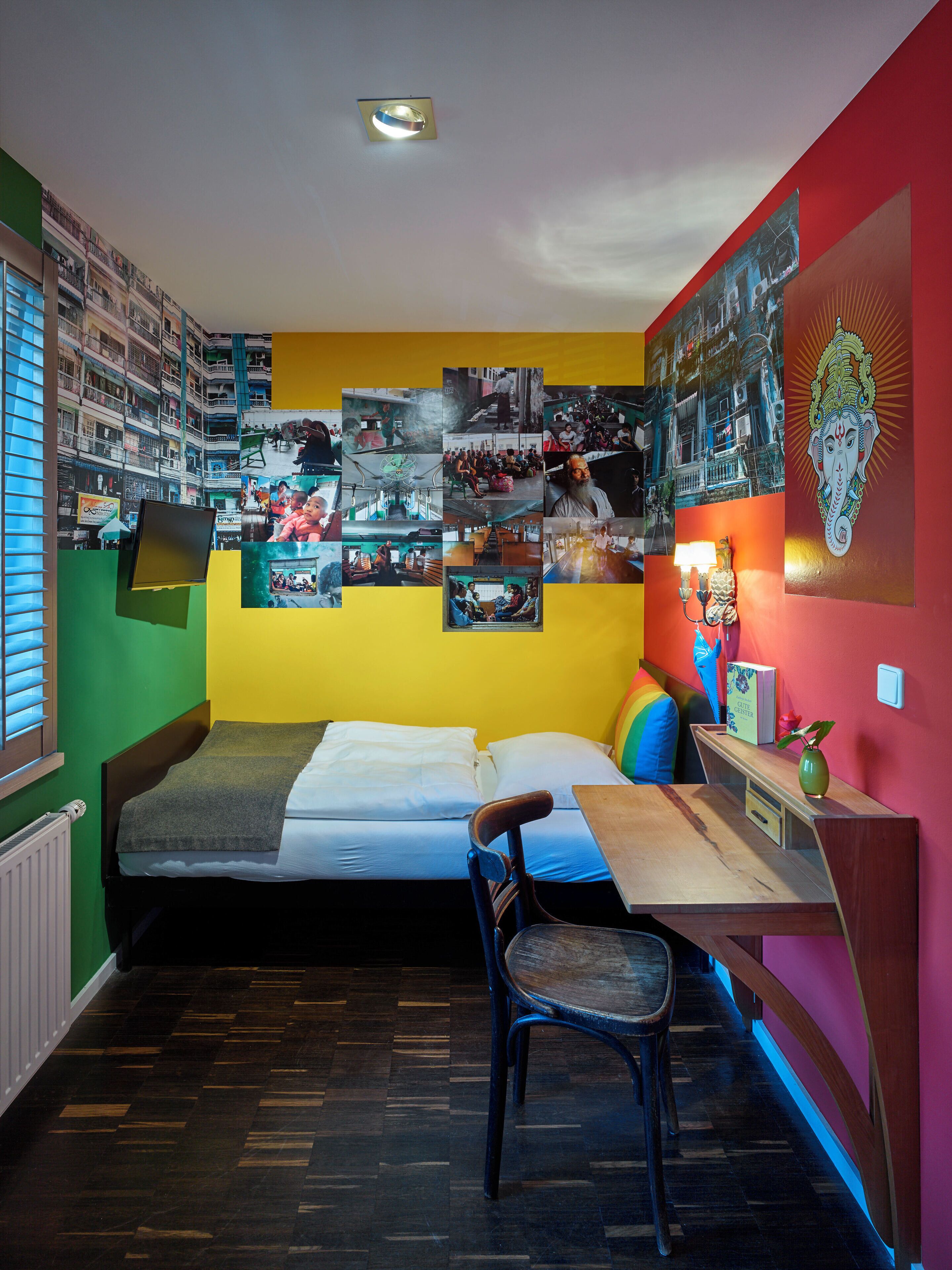 Quarto pequeno e colorido, com uma cama, escrivaninha e cadeira. Há quadros e cartazes nas paredes, e uma janela com persianas.