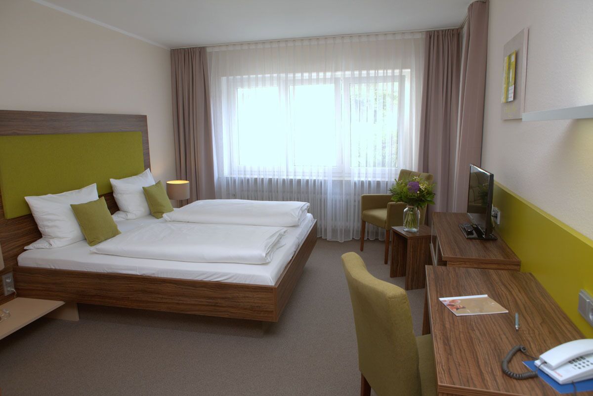 Quarto de hotel com cama de casal, escrivaninha com telefone, cadeira e televisão.