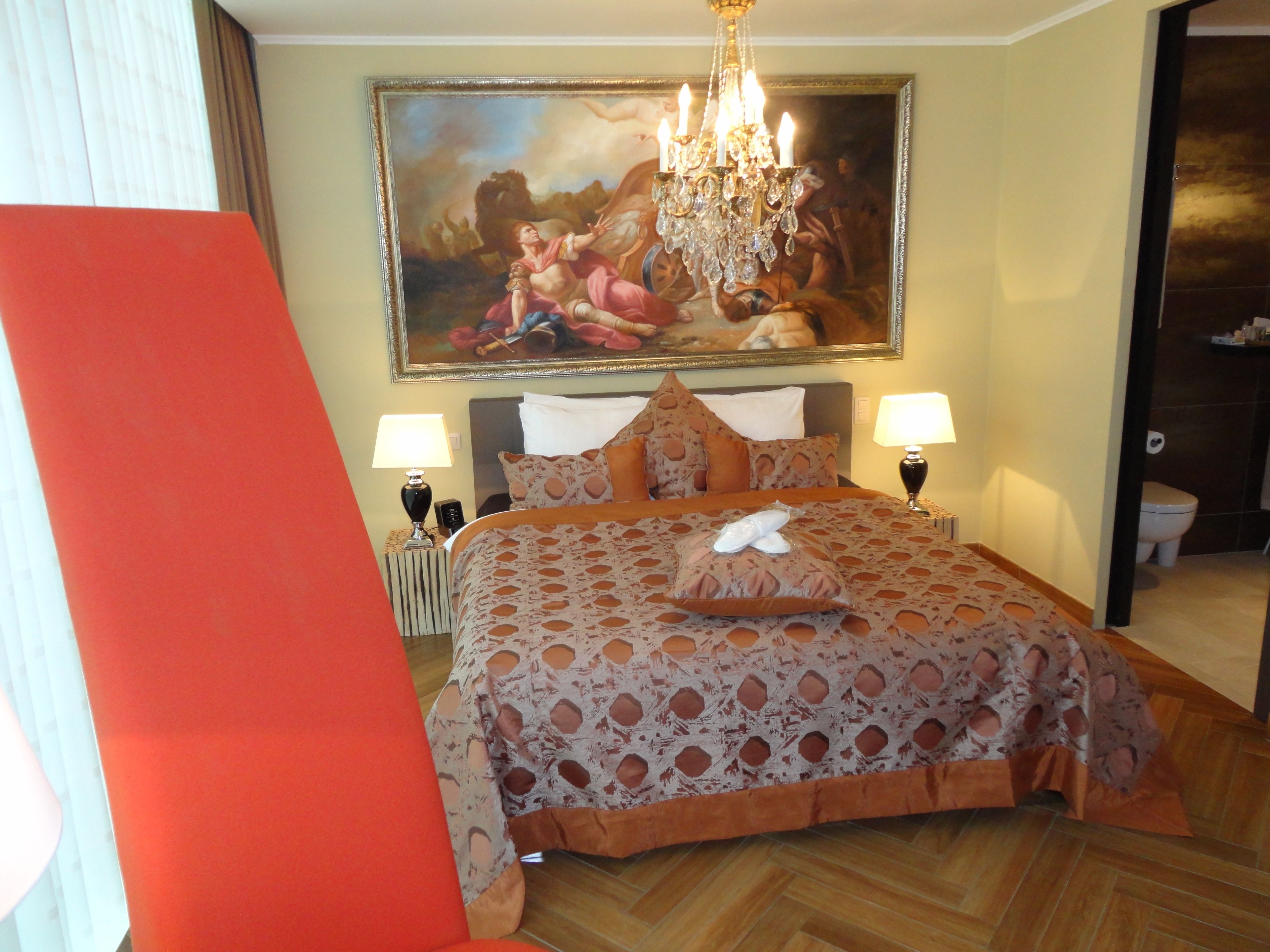 Quarto de hotel com uma cama grande, duas luminárias de cabeceira, um quadro sobre a cama e um candelabro.