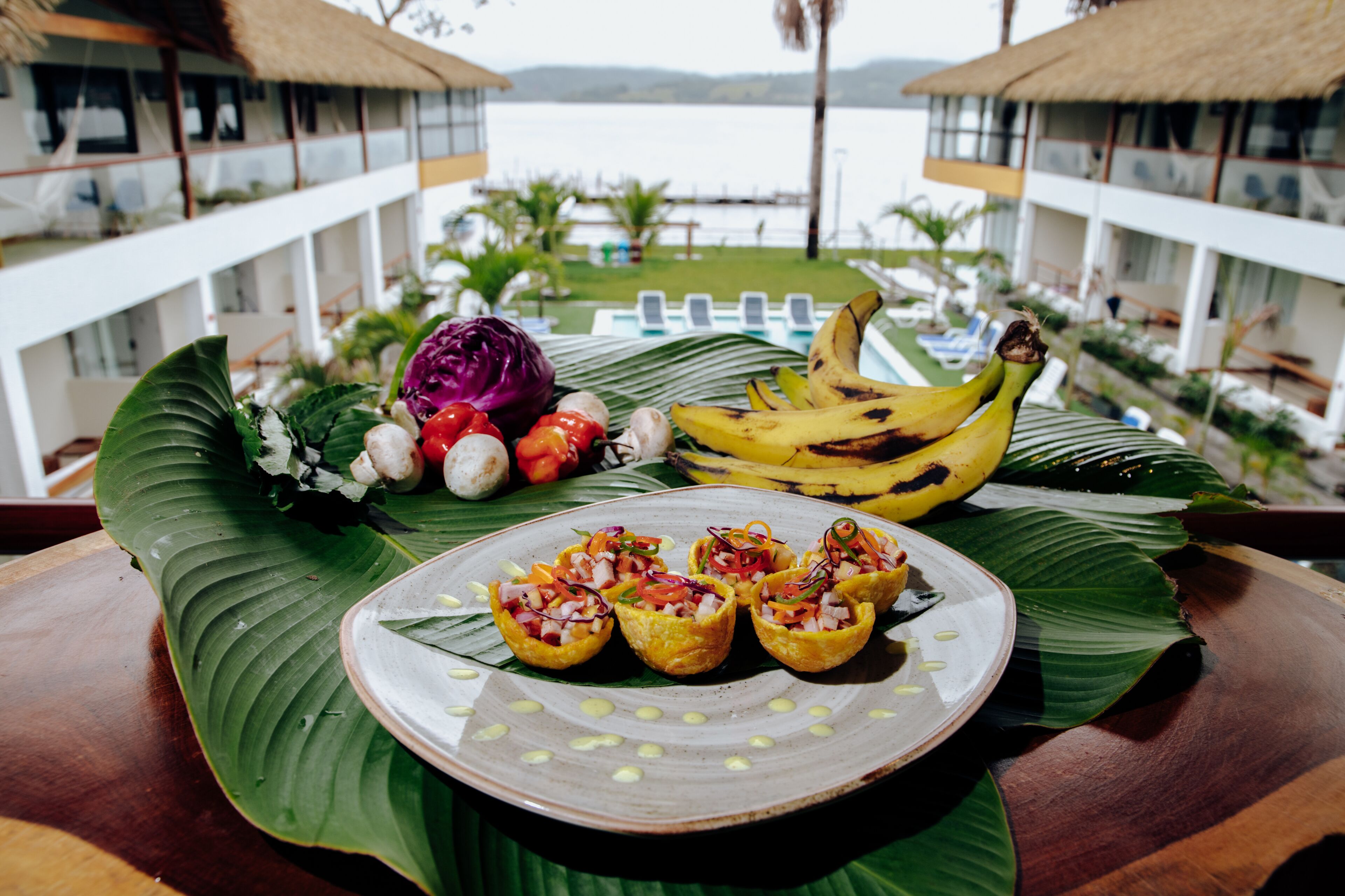 Uma mesa com um prato de aperitivos, uma banana e uma tigela de frutas sobre uma folha.