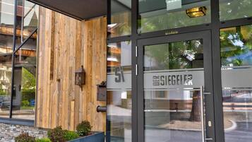 Entrada de um edifício moderno com fachada de madeira, portas de vidro e uma placa com o texto "DER SIEGELEIN".
