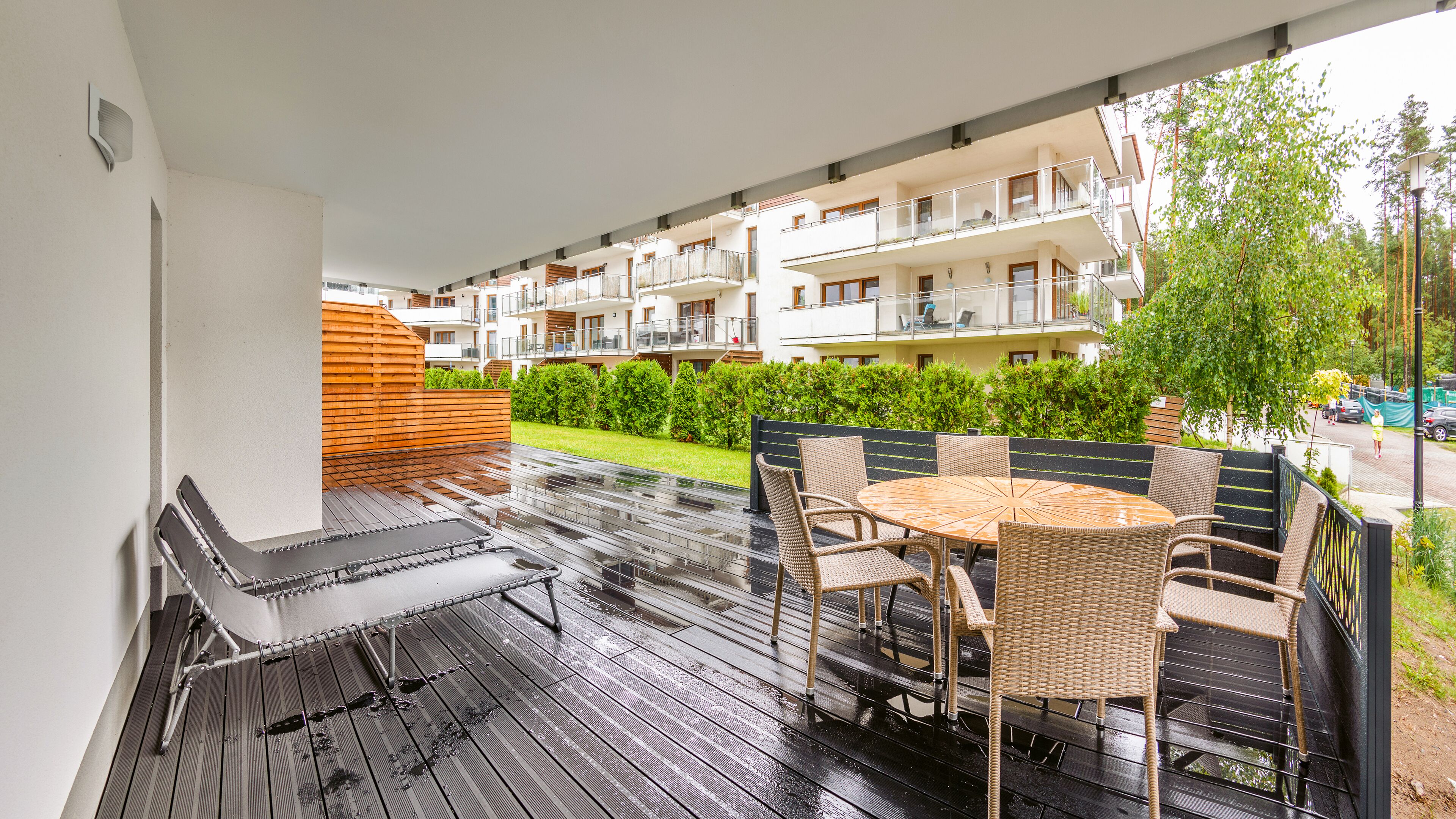 Um alpendre exterior molhado com uma mesa redonda e cadeiras de vime, rodeado por um deck de madeira e vista para um edifício residencial.