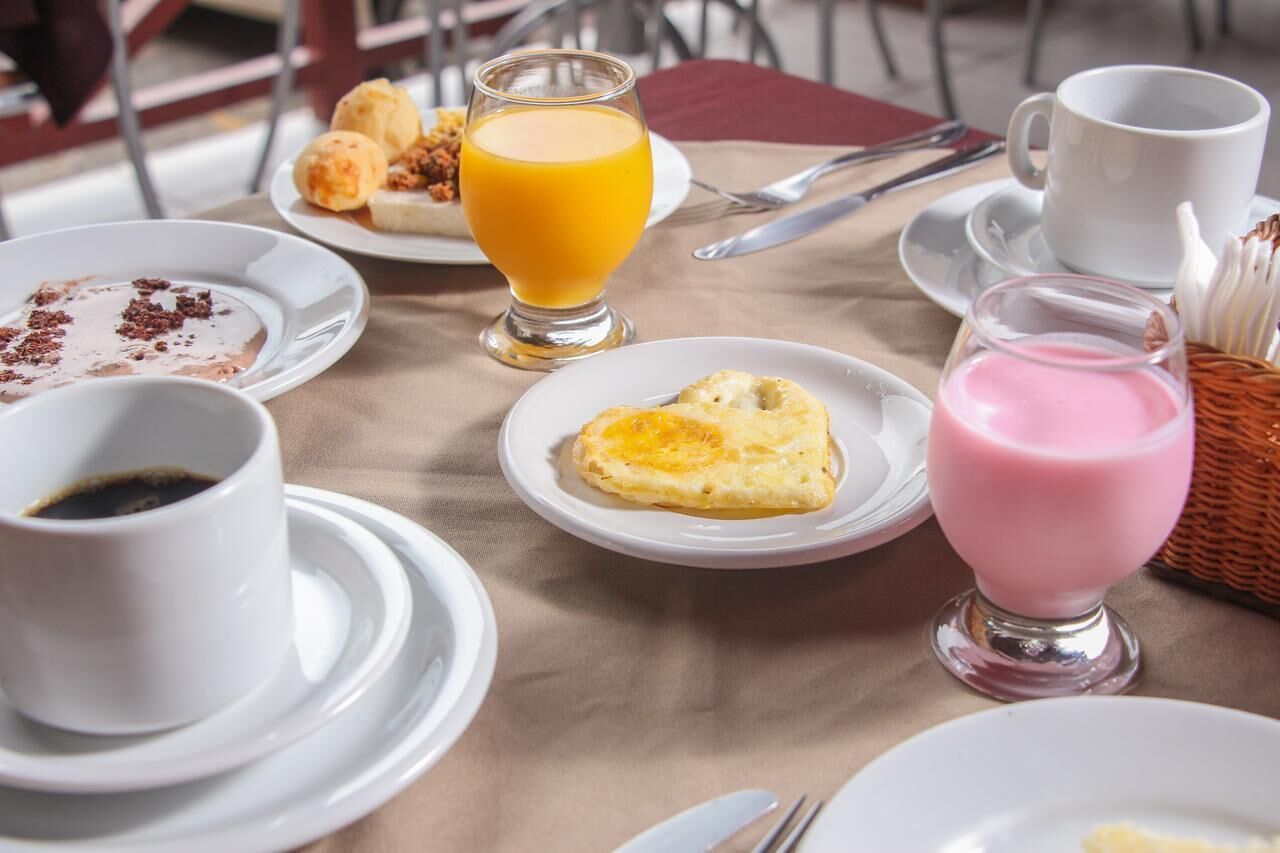 Mesa de café da manhã com pratos de comida, um copo de suco de laranja, uma xícara de café e um copo de bebida rosa.