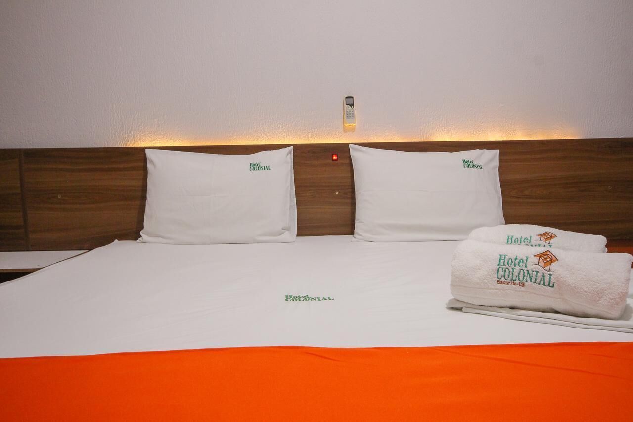 Quarto de hotel com cabeceira de madeira, duas almofadas brancas, cama bem arrumada com colcha laranja e toalha dobrada com o logotipo do hotel.