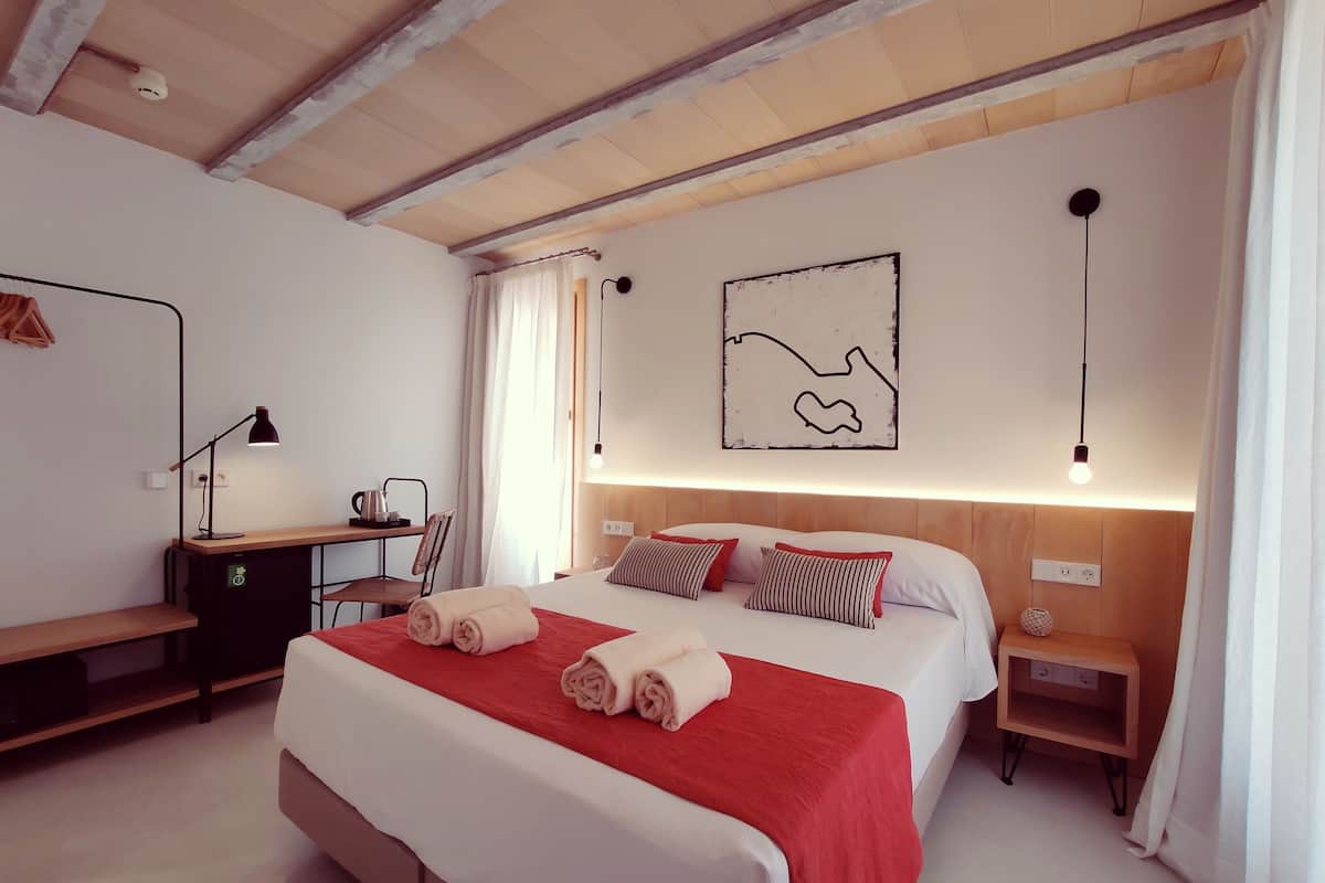 Quarto com cama, mesas de cabeceira, escrivaninha, cadeira, luminária e uma obra de arte na parede.