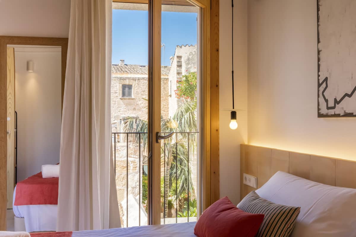 Quarto com varanda, cama com roupa de cama vermelha e branca, e vista para um edifício de pedra e vegetação.