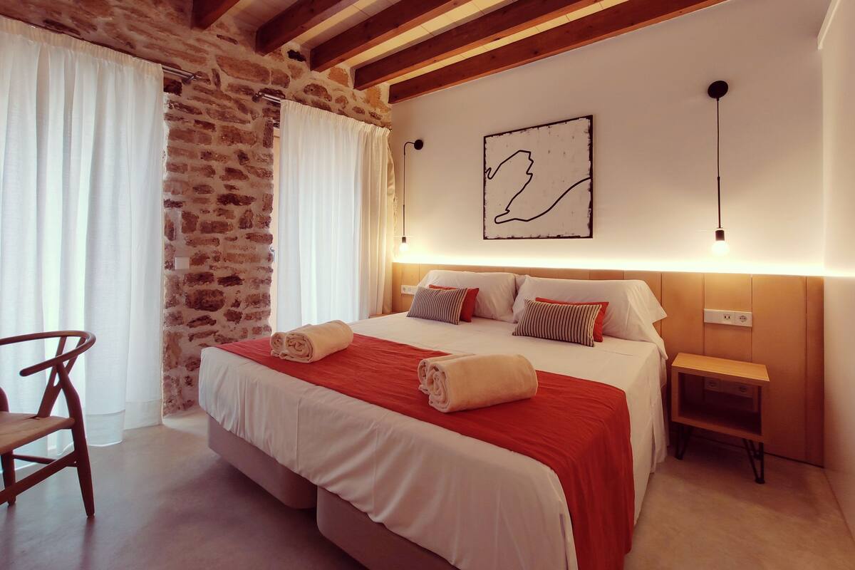 Quarto com parede de pedra, vigas de madeira, cama com roupa de cama vermelha e branca, uma pequena mesa de cabeceira de madeira e uma cadeira.
