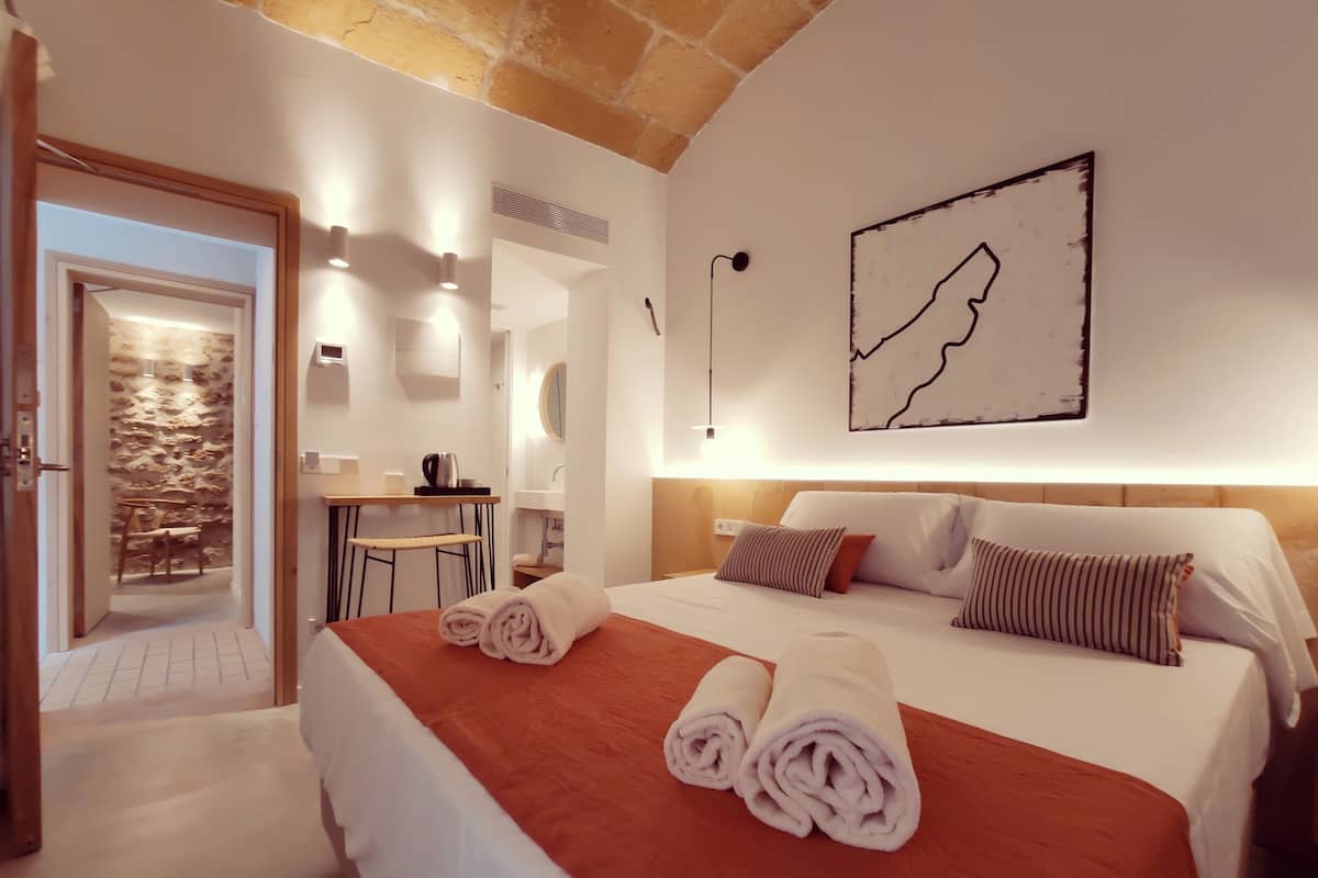Quarto de hotel com uma cama grande, uma escrivaninha com cadeira, uma mesinha e uma parede de pedra.