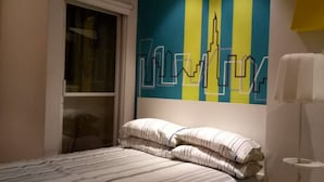 Quarto com cama, almofadas, abajur, ar condicionado e uma parede com arte urbana colorida.