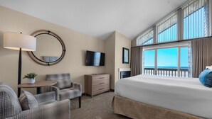 Quarto de hotel com uma cama grande, uma escrivaninha com luminária, uma cadeira, um aparador, uma televisão e vista para o mar.