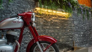 Uma motocicleta vermelha antiga estacionada em frente a uma parede com ripas de madeira verticais e uma placa de neon com a inscrição "Epidamn's Garden".