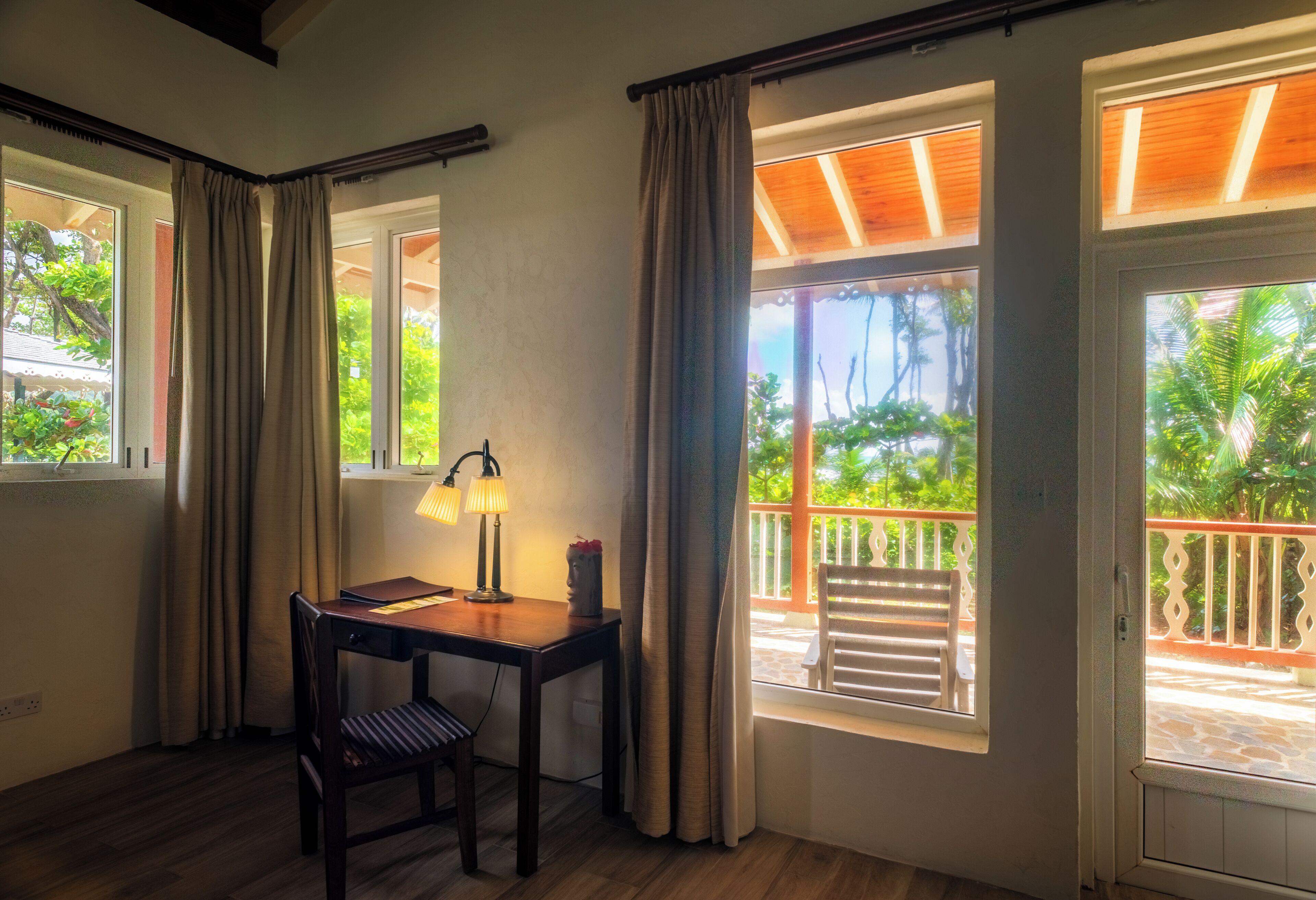 Quarto com escrivaninha de madeira, cadeira e luminária. Há cortinas nas janelas e varanda com grades visível.