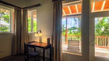 Quarto com escrivaninha de madeira, cadeira e luminária. Há cortinas nas janelas e varanda com grades visível.