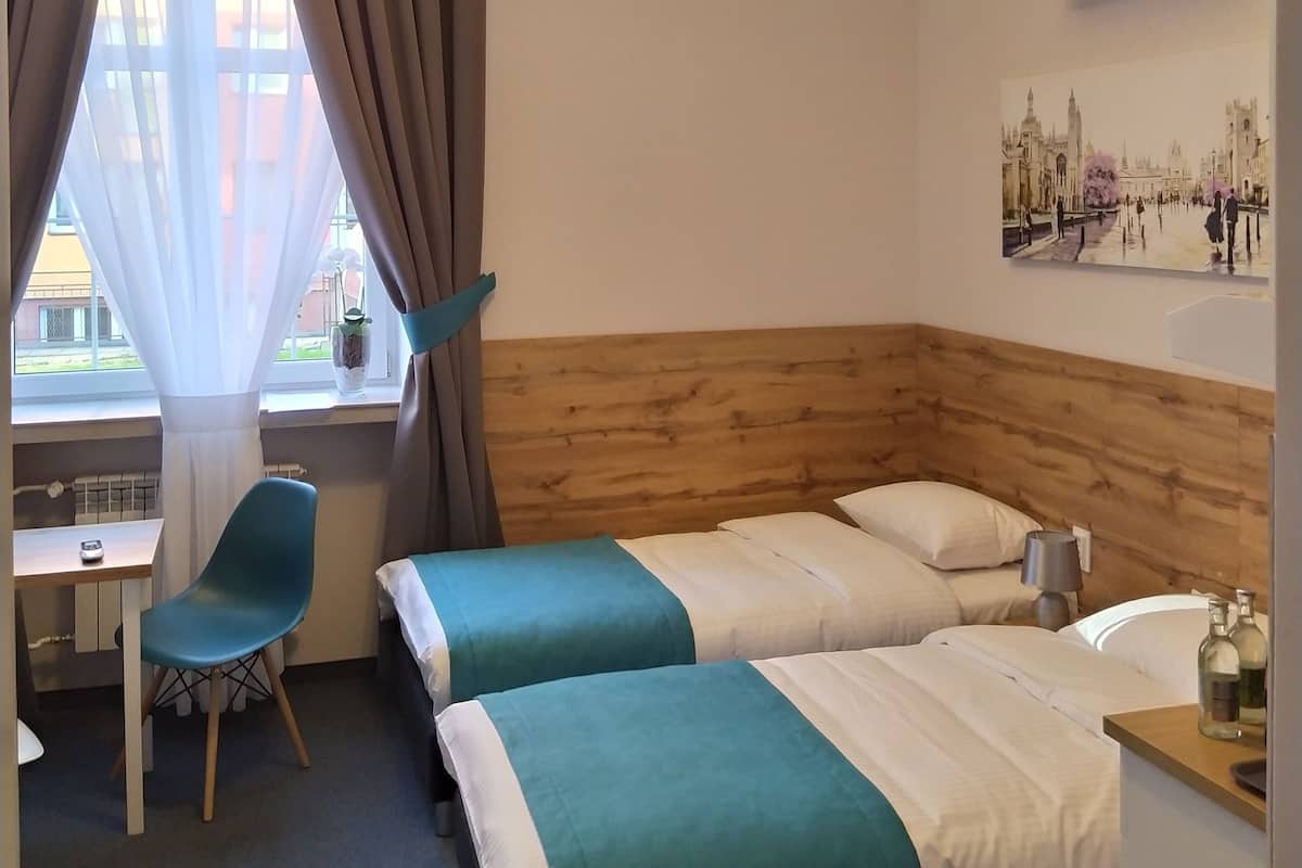 Quarto de hotel com duas camas, uma escrivaninha, uma cadeira e uma janela com cortinas.
