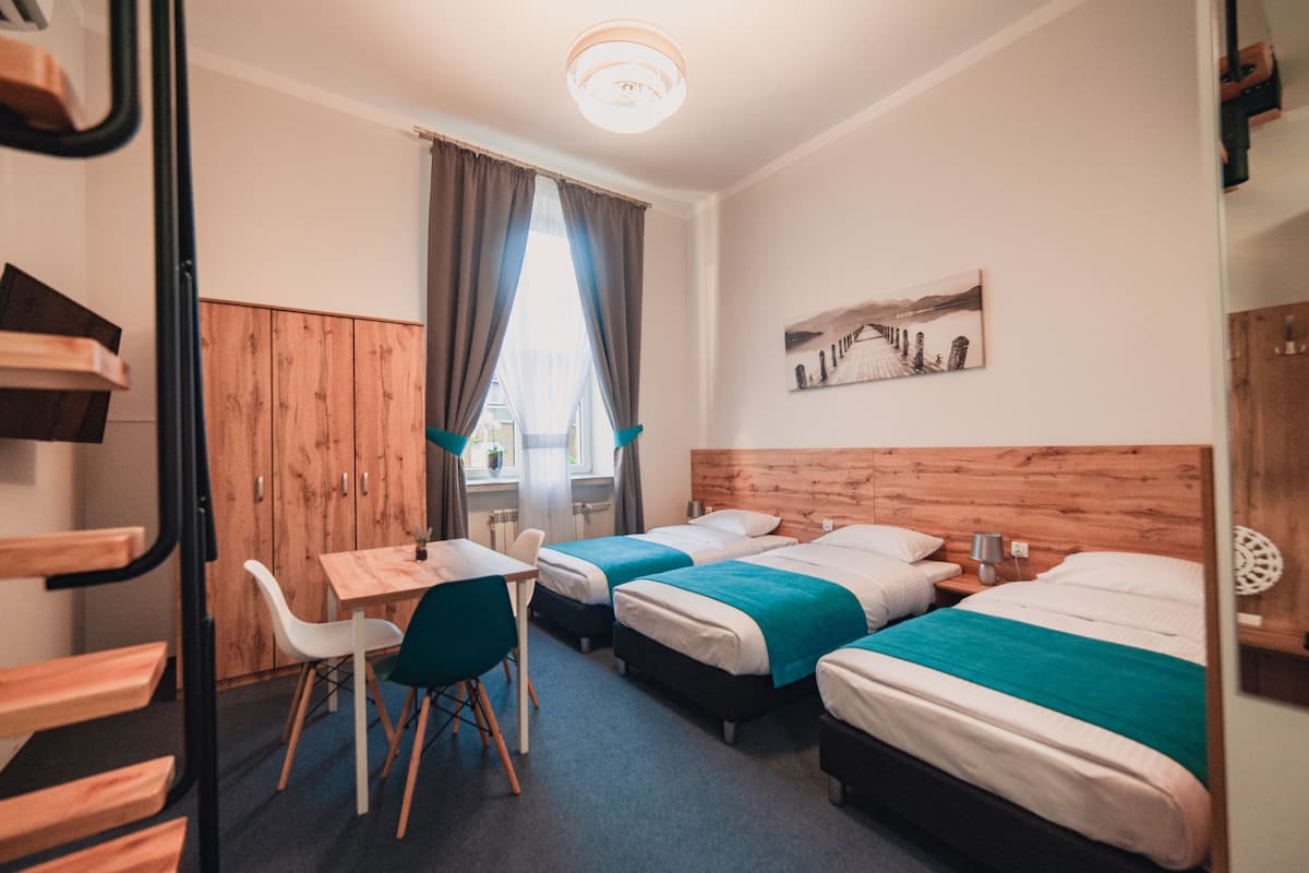 Quarto de hotel com duas camas, cabeceira de madeira, uma mesa com cadeiras e uma janela com cortinas.