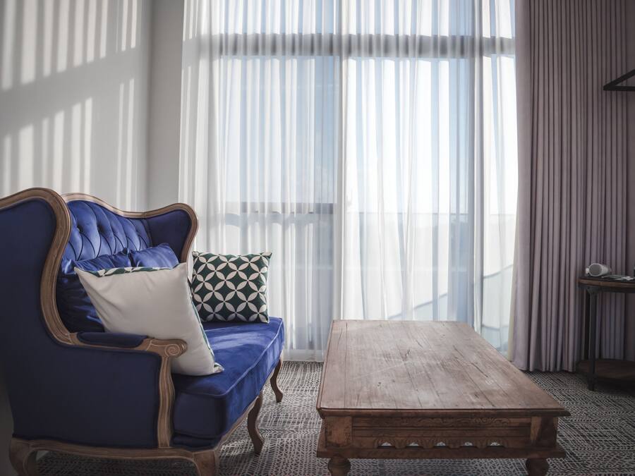 Quarto com sofá azul, mesa de centro de madeira e janela com cortinas leves.