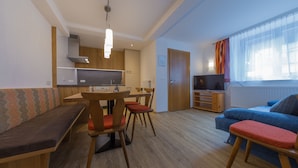 Uma sala de estar moderna com sofá, uma mesa de jantar com cadeiras, televisão e uma cozinha com armários de madeira e forno embutido.