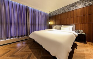 Quarto de hotel com uma cama grande, painéis de madeira nas paredes, mesa de cabeceira com telefone e janela com cortinas.