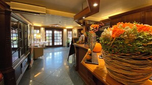 Um saguão de hotel com uma mesa de recepção, um vaso grande de flores e vista para o exterior através de grandes janelas.