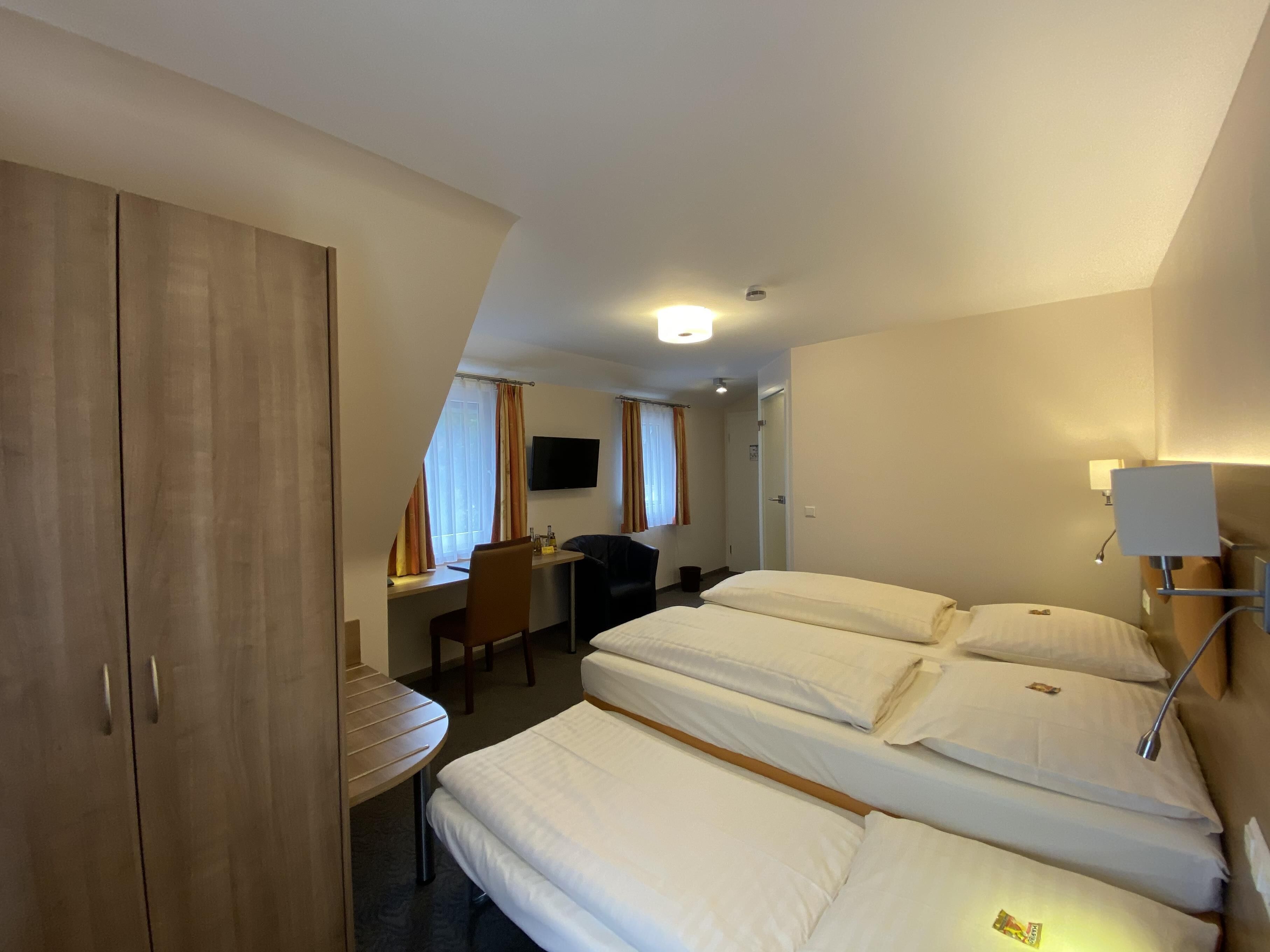 Quarto de hotel com três camas, uma escrivaninha, uma cadeira e uma televisão.
