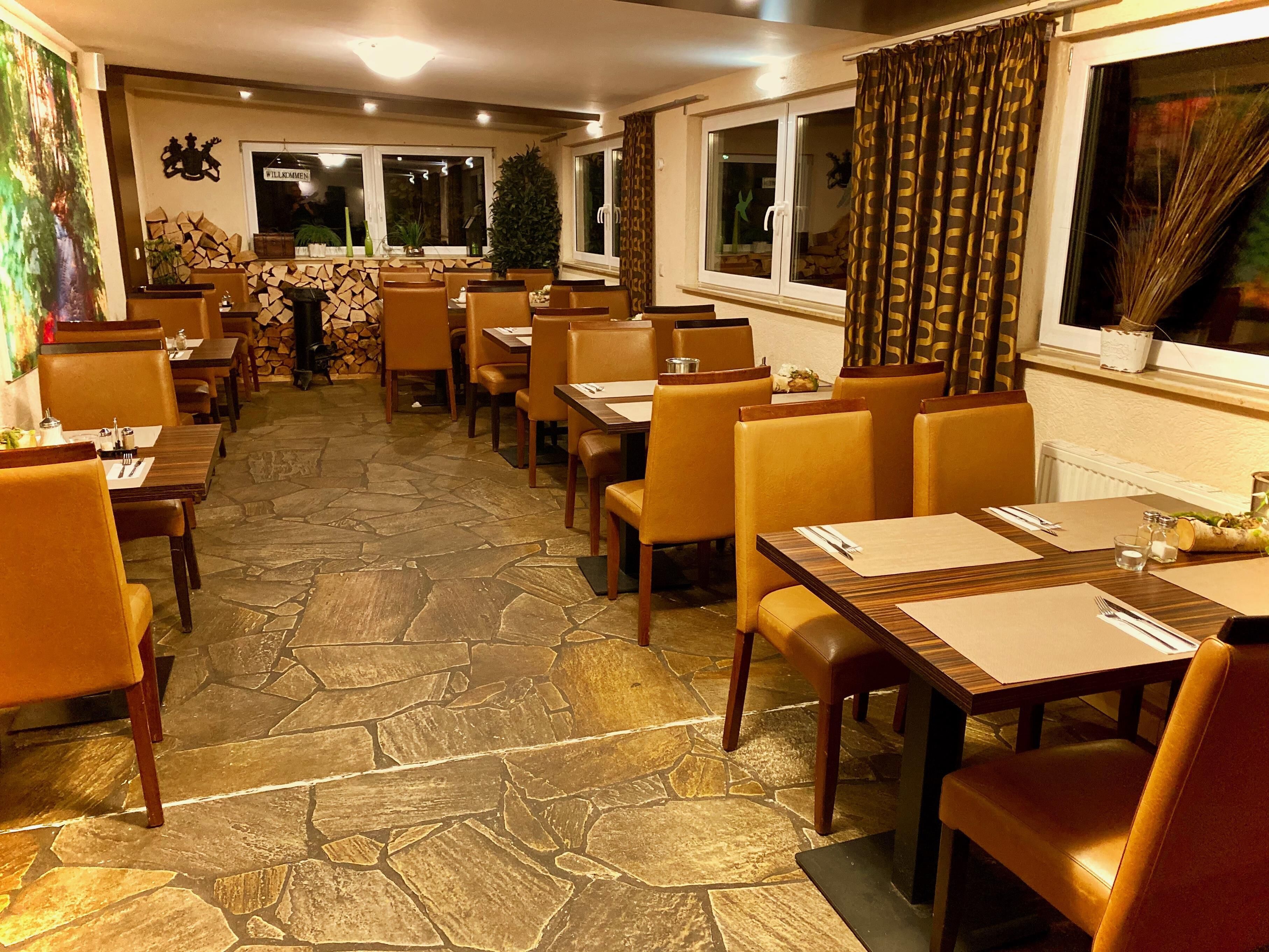 Um restaurante com soalho de pedra, mesas e cadeiras de madeira e grandes janelas com cortinas.