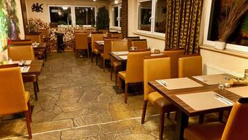 Um restaurante com soalho de pedra, mesas e cadeiras de madeira e grandes janelas com cortinas.