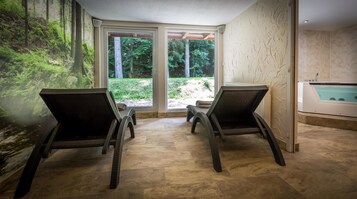 Um quarto de spa com duas poltronas de descanso pretas, uma janela grande com vista para uma floresta e uma banheira branca.