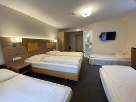 Quarto de hotel com três camas de solteiro, cabeceira de madeira, abajures, televisão de tela plana e porta que dá para um banheiro.