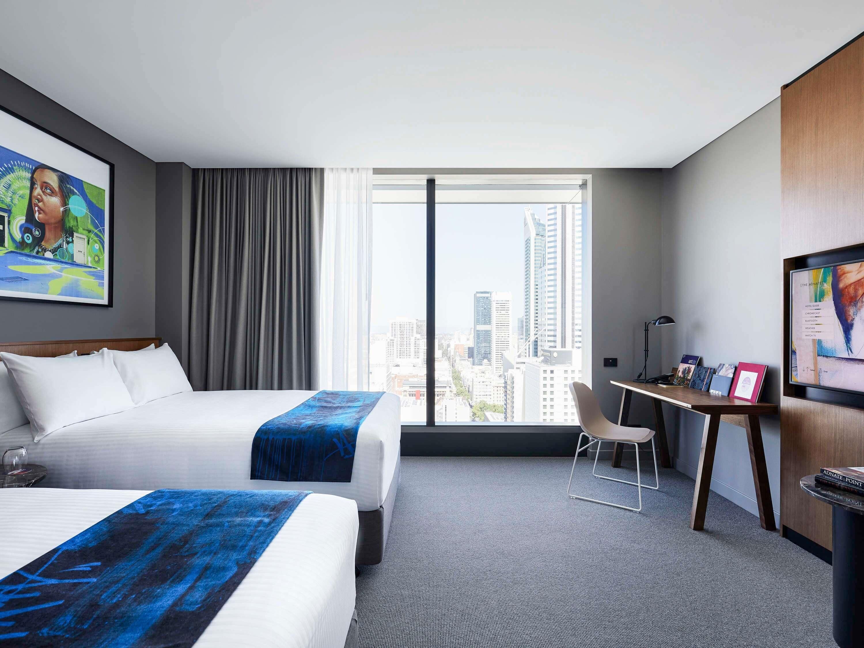 Quarto de hotel com duas camas, uma escrivaninha, uma cadeira, uma janela grande com vista para a cidade e um quadro na parede.