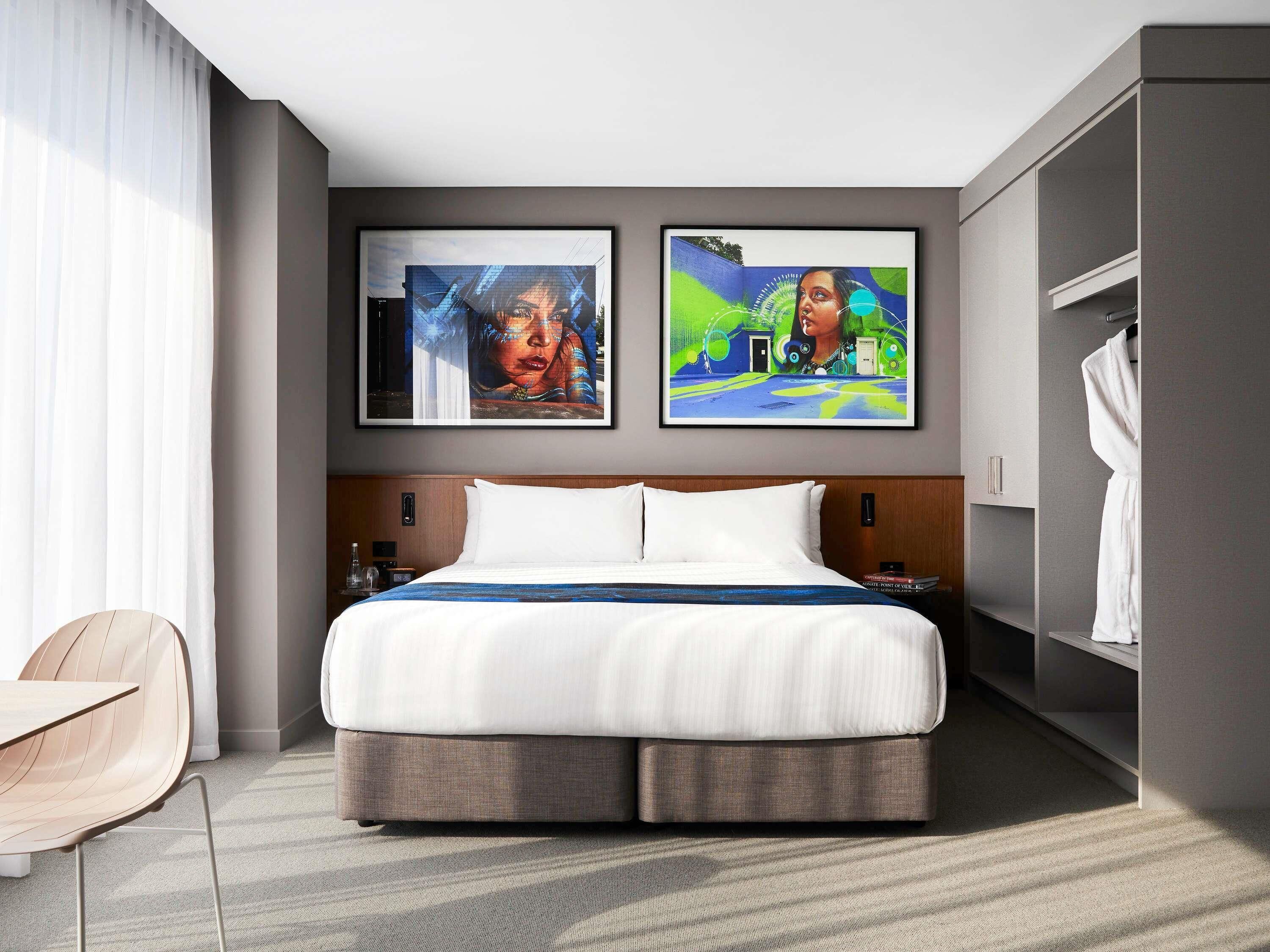 Quarto de hotel moderno com uma cama grande, uma cadeira de madeira, um guarda-roupa e duas obras de arte emolduradas na parede.