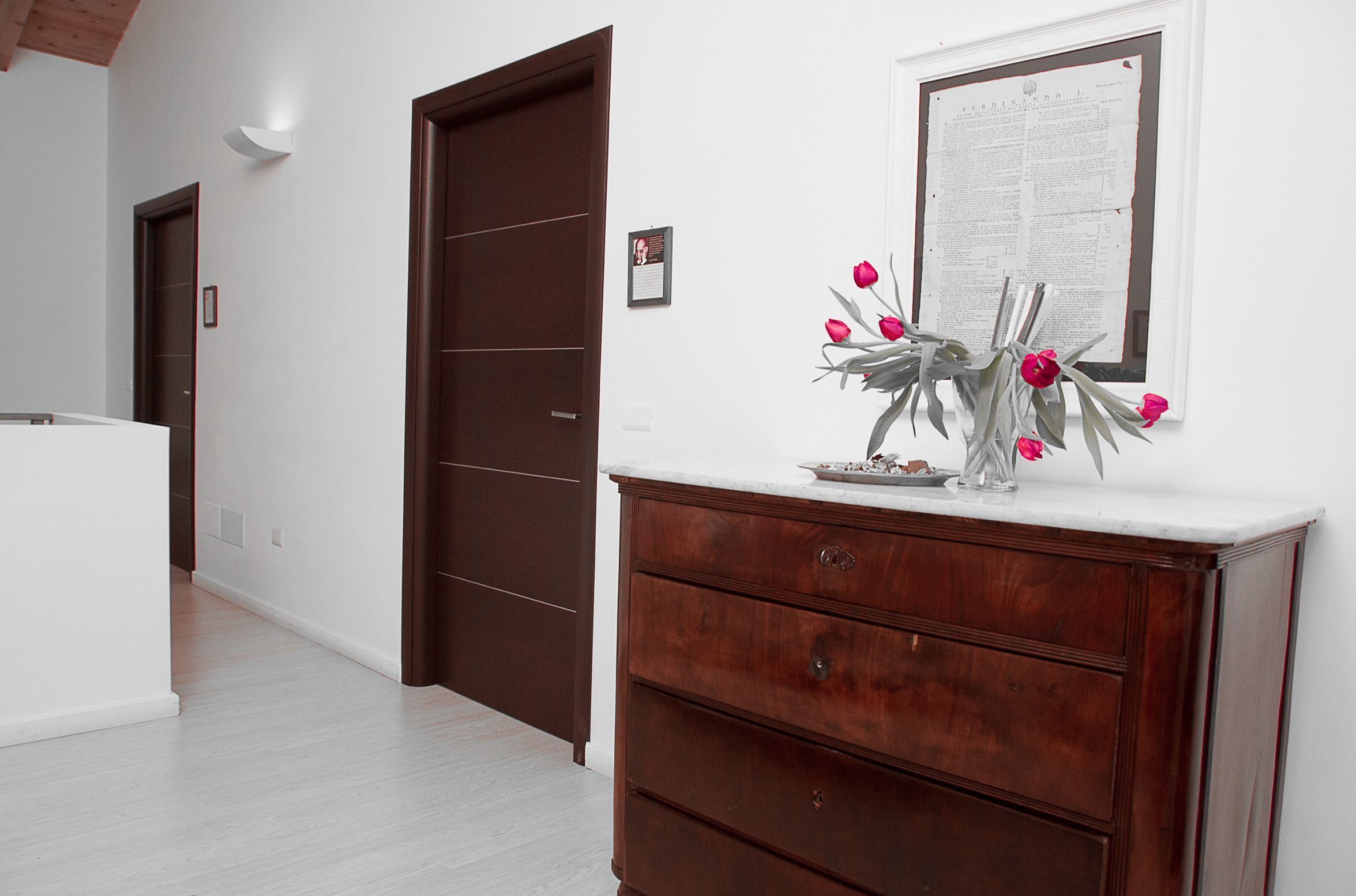 Um corredor com porta de madeira, parede branca com documento emoldurado e cômoda de madeira com um vaso de flores.