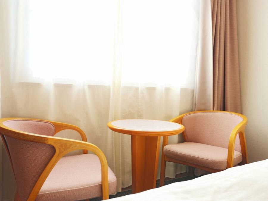 Quarto de hotel com uma cama, duas cadeiras de madeira com almofadas cor-de-rosa e uma mesa redonda pequena.