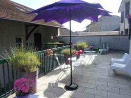Uma área de pátio com um guarda-sol roxo, plantas em vasos e mesas e cadeiras ao ar livre.
