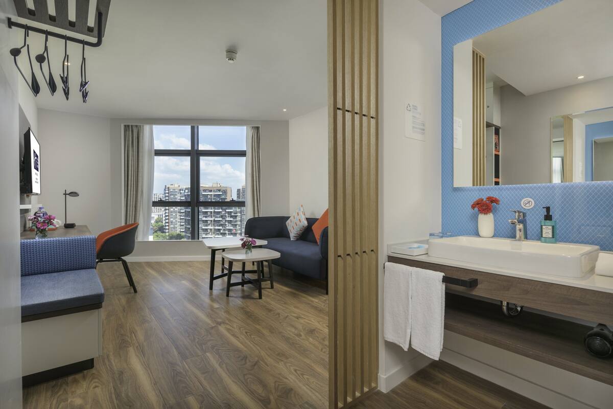 Quarto de hotel moderno com uma janela grande, um sofá, uma mesa pequena, banheiro com pia e espelho, e vista para a cidade.