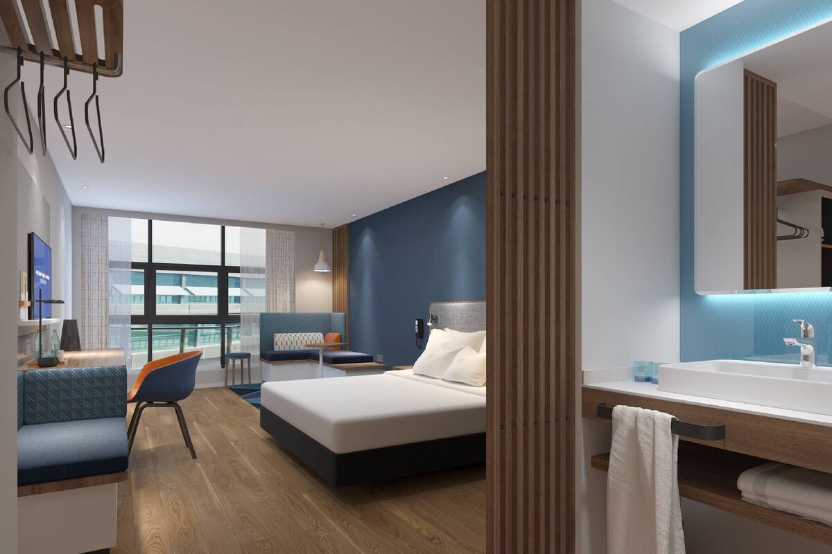 Quarto de hotel moderno com uma cama grande, área de estar com uma cadeira e mesa, uma janela ampla e banheiro com pia e espelho.