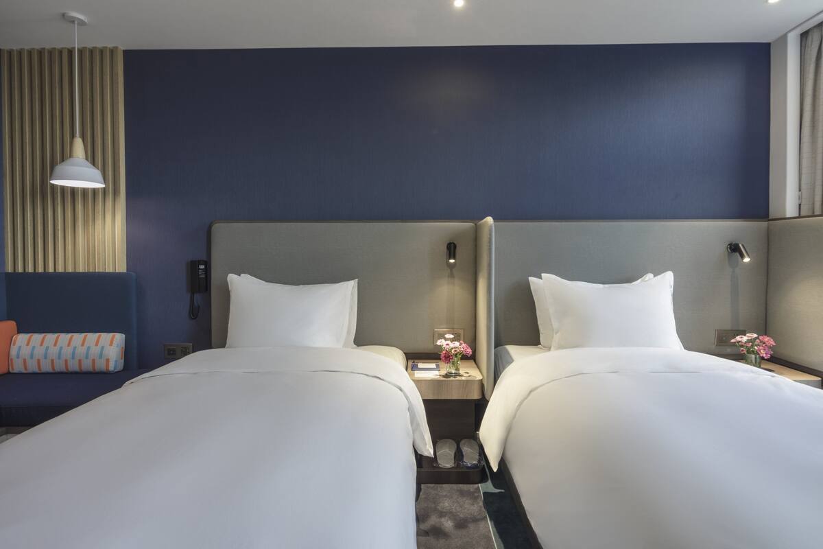 Quarto de hotel com duas camas, uma mesa de cabeceira com abajur, telefone e um pequeno vaso com flores.