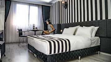 Quarto de hotel com uma cama grande, uma mesa pequena com uma garrafa e copos, uma cadeira e uma escrivaninha.