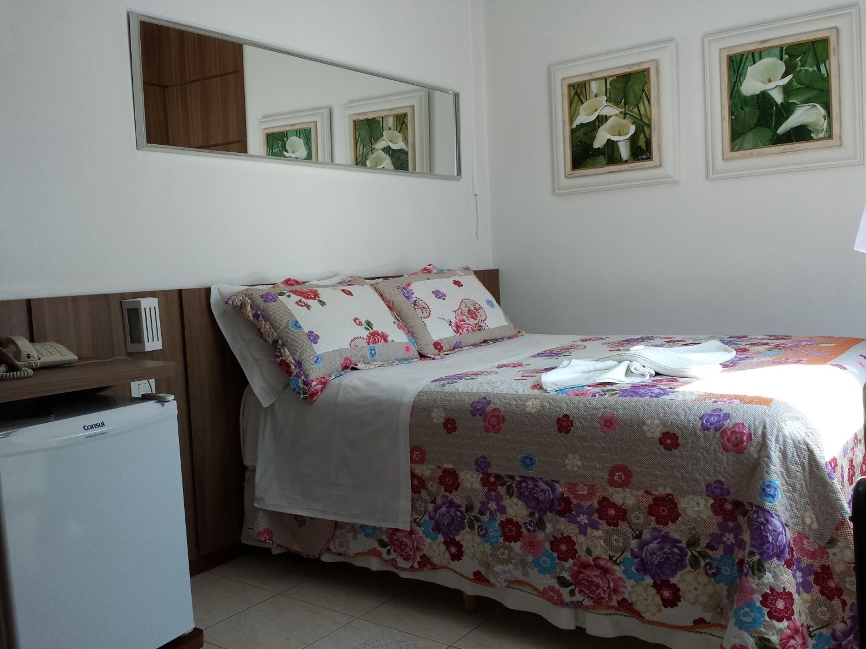 Quarto com cama, mesa de cabeceira com telefone e pequeno frigorífico.