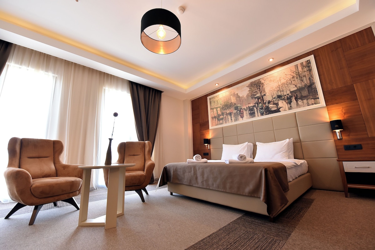 Quarto de hotel com uma cama, duas poltronas, uma mesa pequena, um quadro grande na parede e uma mesa de cabeceira com abajur.