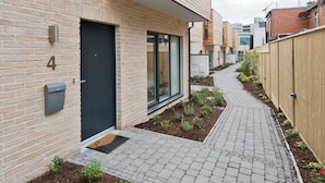 Um edifício residencial moderno com fachada de tijolos, porta de cor escura e um pequeno jardim com plantas e um caminho.