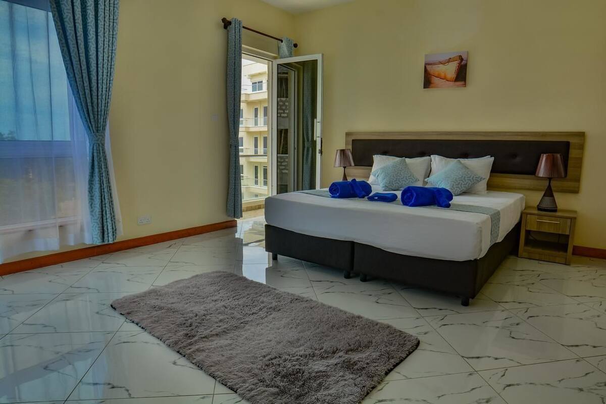 Quarto de hotel com uma cama grande, um tapete macio, uma mesinha de cabeceira e uma janela com cortinas.