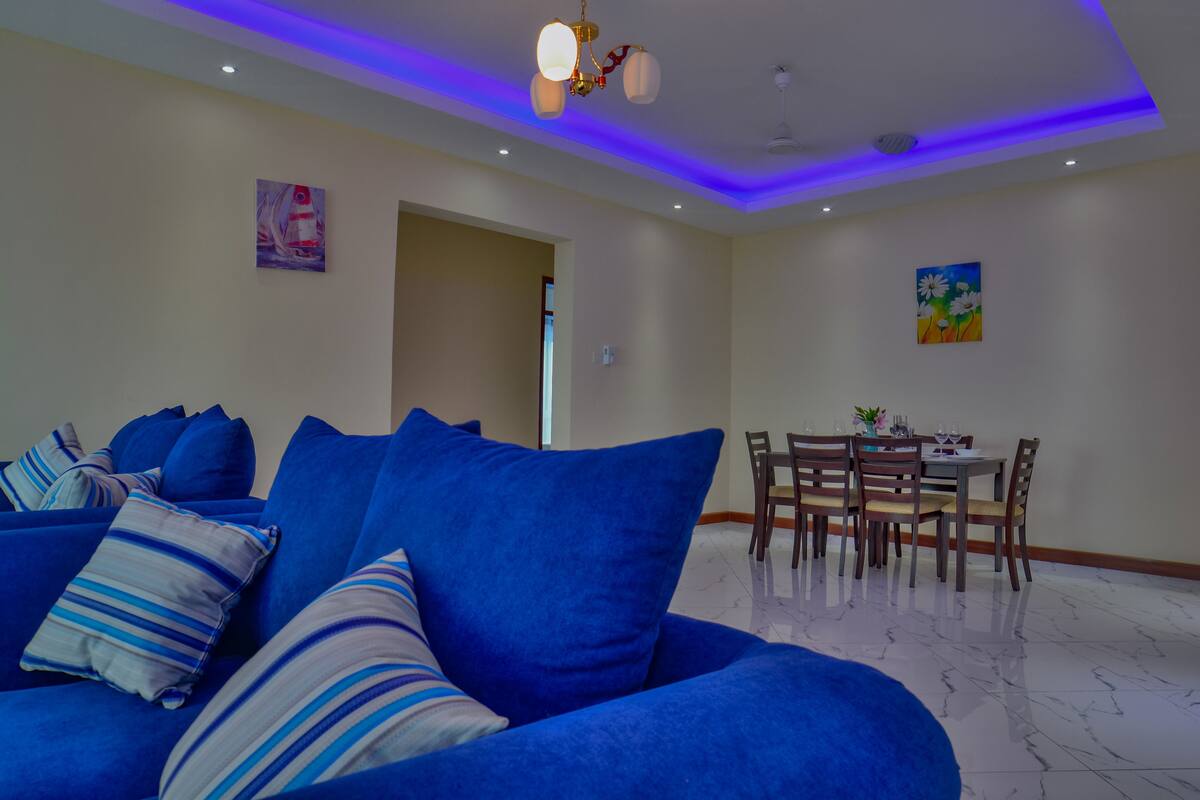 Uma sala de estar com um sofá azul, uma mesa de jantar com cadeiras e um teto com iluminação azul.