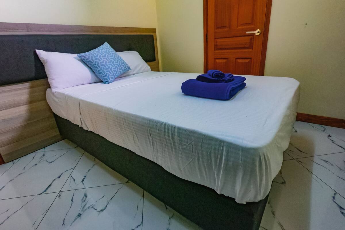 Cama bem arrumada com colcha branca e toalha azul, cabeceira de madeira e porta de madeira fechada.
