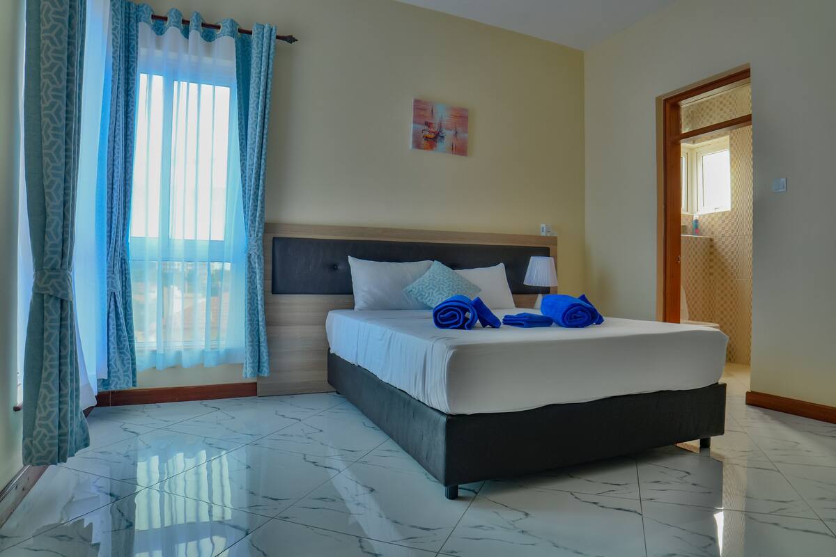 Cama bem arrumada com almofadas e toalhas dobradas, abajur e janela com cortinas estampadas.