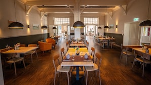 Uma sala de jantar ampla com várias mesas e cadeiras, grandes janelas e iluminação pendente.