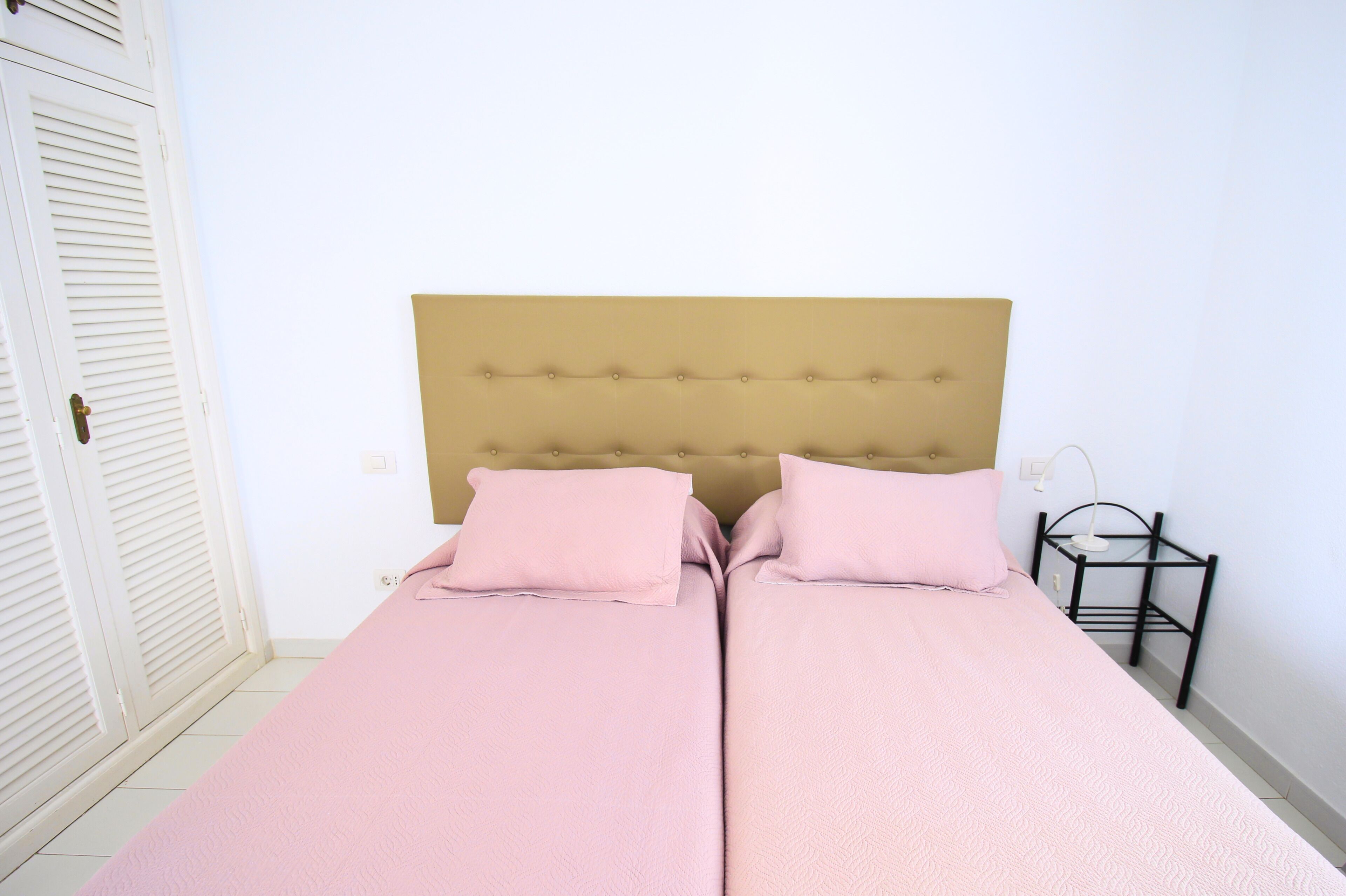 Uma cama de casal com cabeceira, duas almofadas, mesa de cabeceira e luminária na parede.