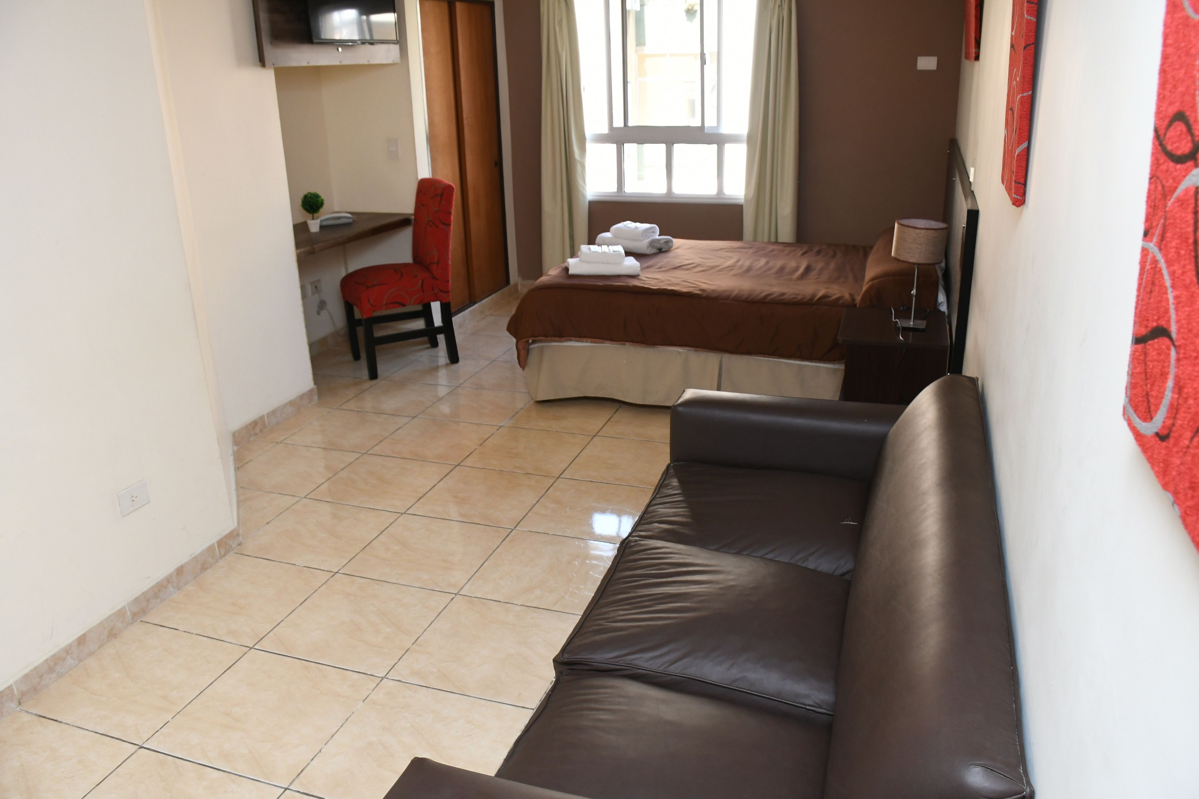 Quarto de hotel com uma cama, uma cadeira vermelha, um sofá marrom, uma televisão e uma janela com cortinas.