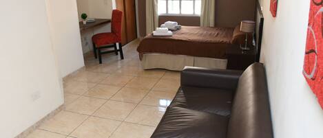 Quarto de hotel com uma cama, uma cadeira vermelha, um sofá marrom, uma televisão e uma janela com cortinas.