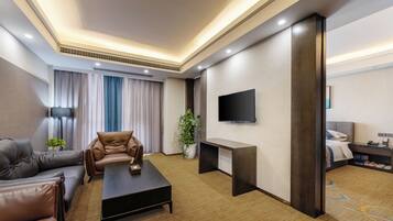 Quarto de hotel moderno com sofá de couro, mesa de centro, televisão fixada na parede e uma pequena mesa de cabeceira com abajur e telefone.